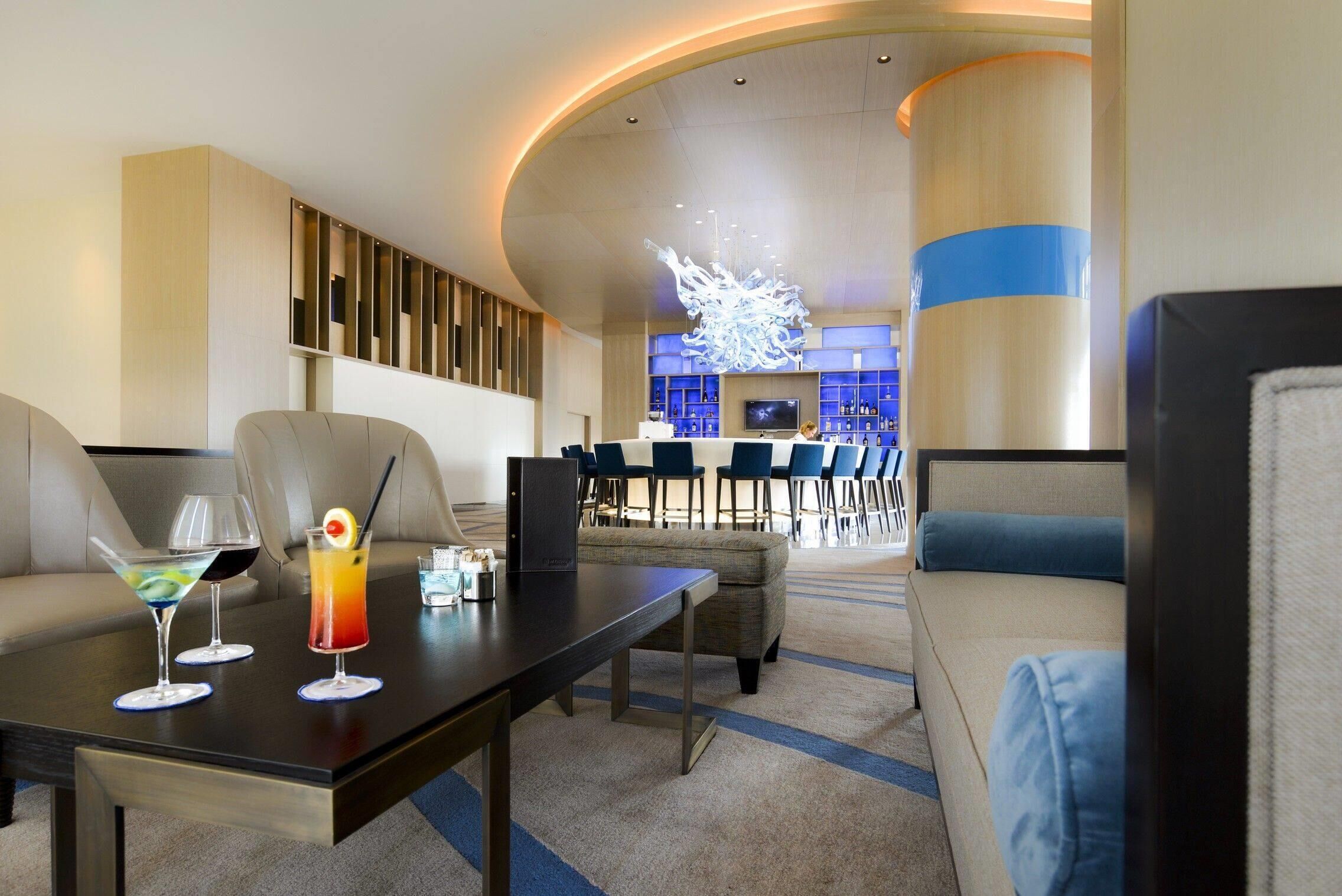 Um saguão de hotel moderno com área de bar, espaços de estar e um detalhe decorativo no teto.