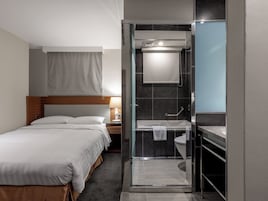 Quarto de hotel com cama, mesa de cabeceira, luminária e chuveiro em gabinete de vidro, pia e vaso sanitário.