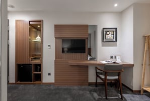 Quarto de hotel compacto com escrivaninha integrada, televisão, uma cadeira e guarda-roupa com espelho.