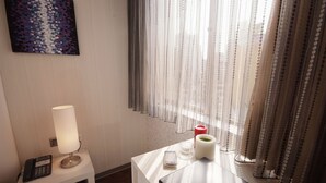 Quarto de hotel com escrivaninha, luminária, telefone, controle remoto e janela com cortinas leves.