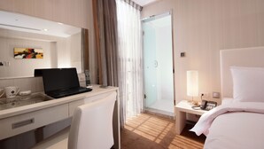 Quarto de hotel com escrivaninha, laptop, cadeira, cama, mesa de cabeceira, abajur e divisória de chuveiro de vidro.