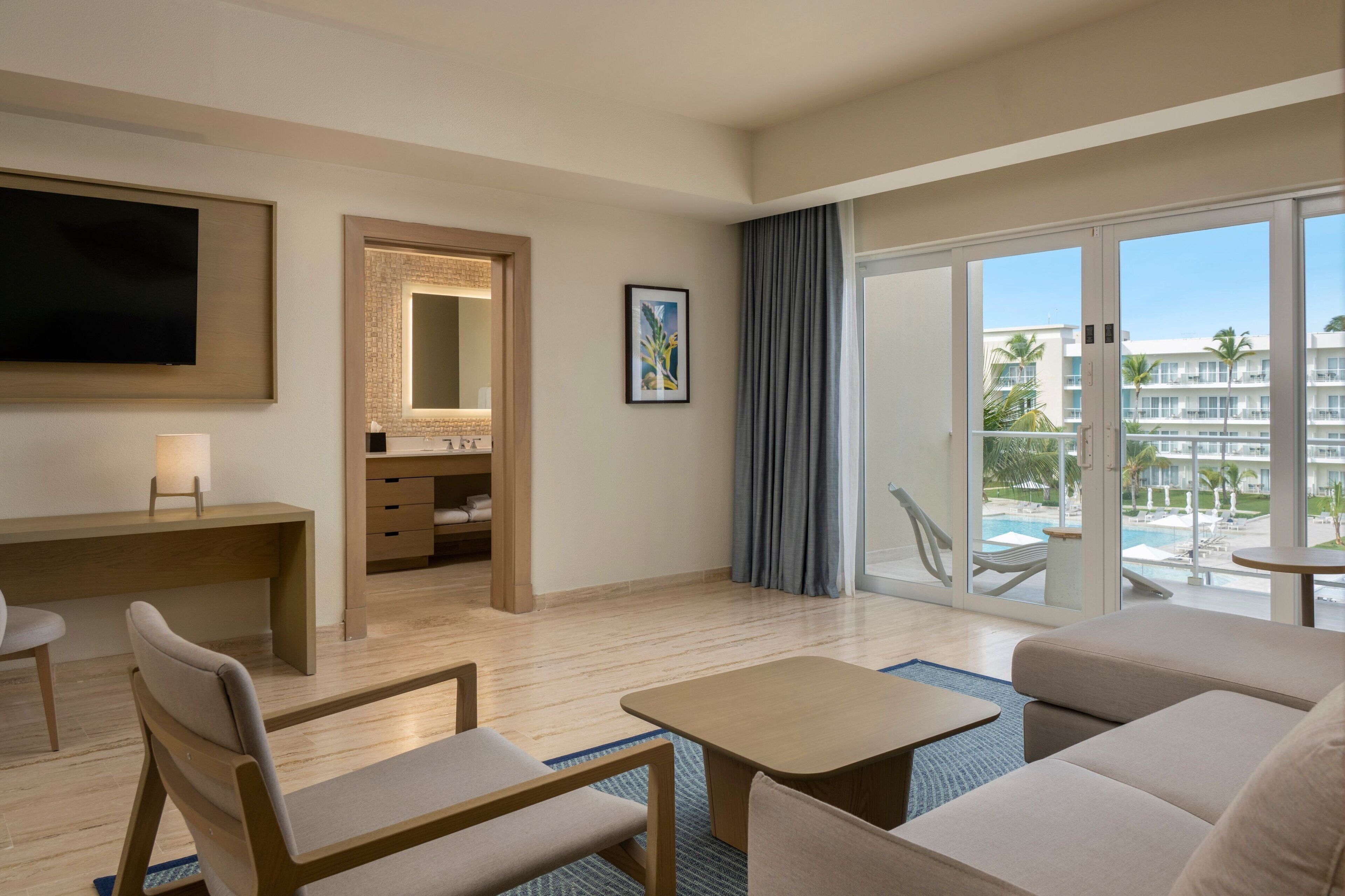 Quarto de hotel com televisão, escrivaninha, sofá, mesinha, espelho, quadro na parede e vista para a piscina e edifícios externos.