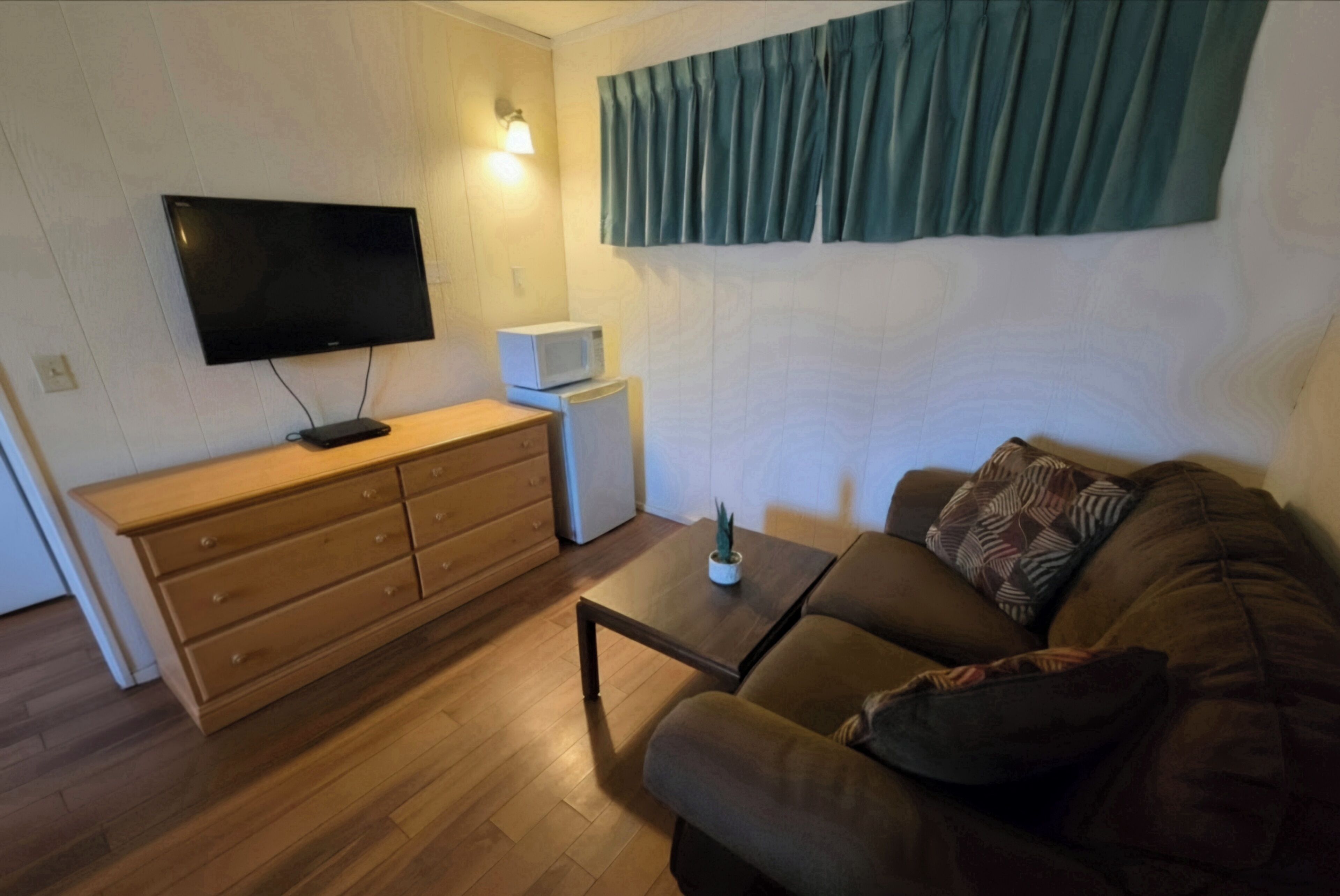 Sala de estar com televisão de tela plana na parede, cômoda de madeira, micro-ondas branco, sofá marrom com almofadas estampadas e uma pequena mesa com uma planta.