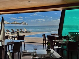 Uma sala de jantar com vista para o oceano, com mesas, cadeiras e guarda-sóis.