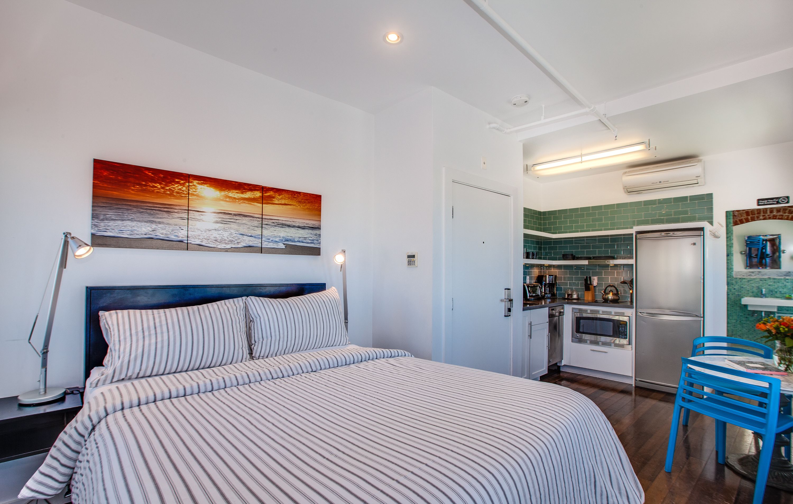 Quarto com cama, mesa de cabeceira, luminária, pintura de um pôr do sol, cozinha compacta com geladeira, micro-ondas e fogão, e uma cadeira azul.