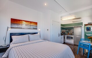 Quarto com cama, mesa de cabeceira, luminária, pintura de um pôr do sol, cozinha compacta com geladeira, micro-ondas e fogão, e uma cadeira azul.