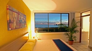 Quarto de hotel com uma grande janela com vista para o mar e um pôr do sol.