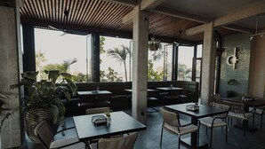 Um espaço de refeições interno com mesas, cadeiras, janelas amplas e plantas em vasos.