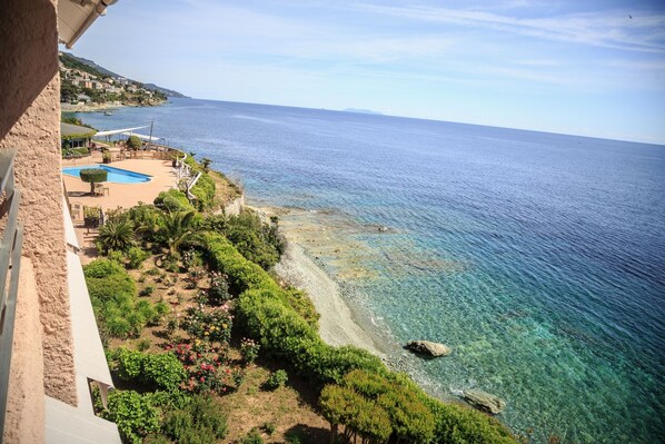 Vista costeira com mar azul cristalino, areal arenoso e jardim ajardinado com diversas plantas e flores.