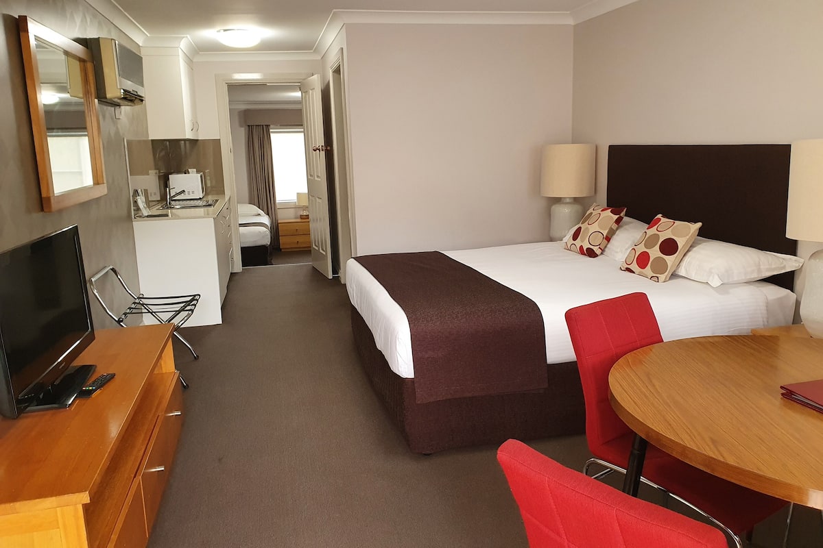 Quarto de hotel com cama, escrivaninha com televisão, cadeira, luminária e mesinha.