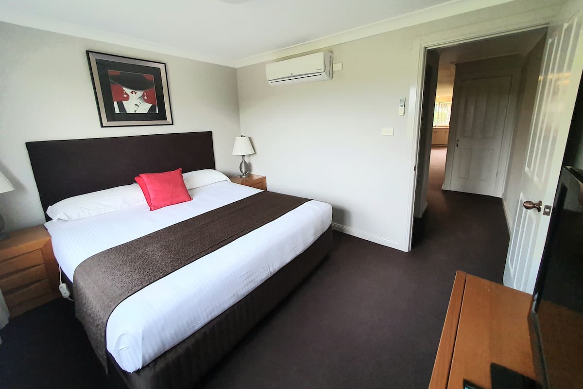Quarto de hotel com uma cama grande, mesa de cabeceira com abajur, aparelho de ar condicionado e porta para outro cômodo.