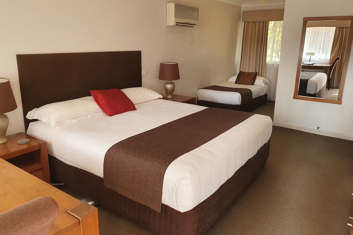 Quarto de hotel com uma cama grande, uma cama menor, uma escrivaninha, uma cadeira, um abajur, um espelho e um aparelho de ar condicionado.