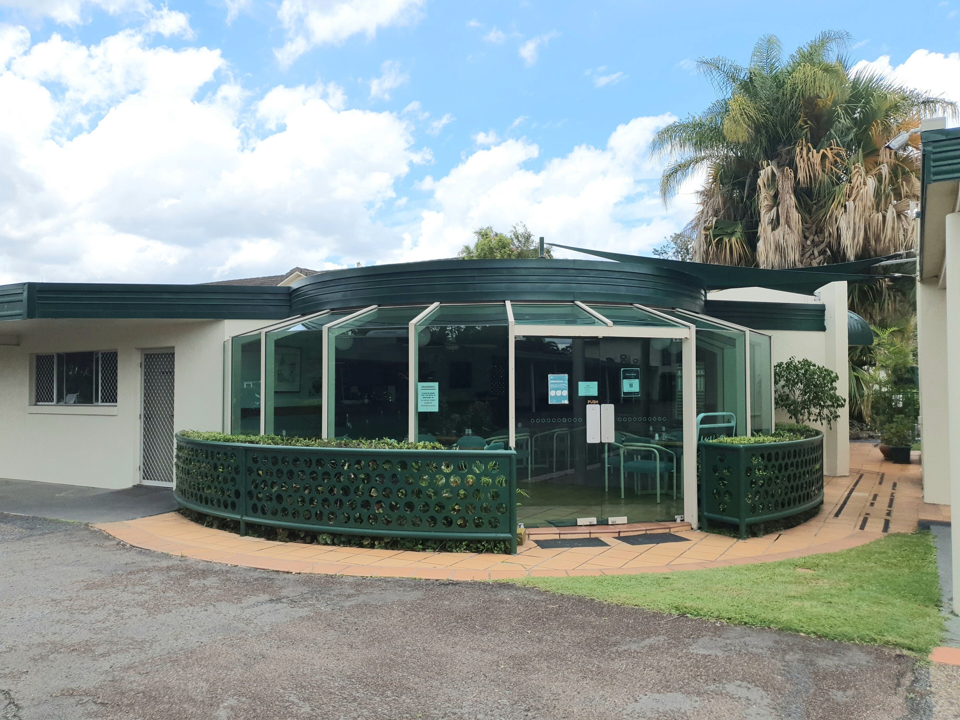 Um edifício com entrada de vidro, telhado verde e uma palmeira ao fundo.