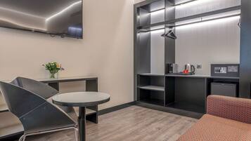 Quarto de hotel moderno com televisão de tela plana, uma pequena mesa com cadeira, sofá, máquina de café e mini-geladeira.