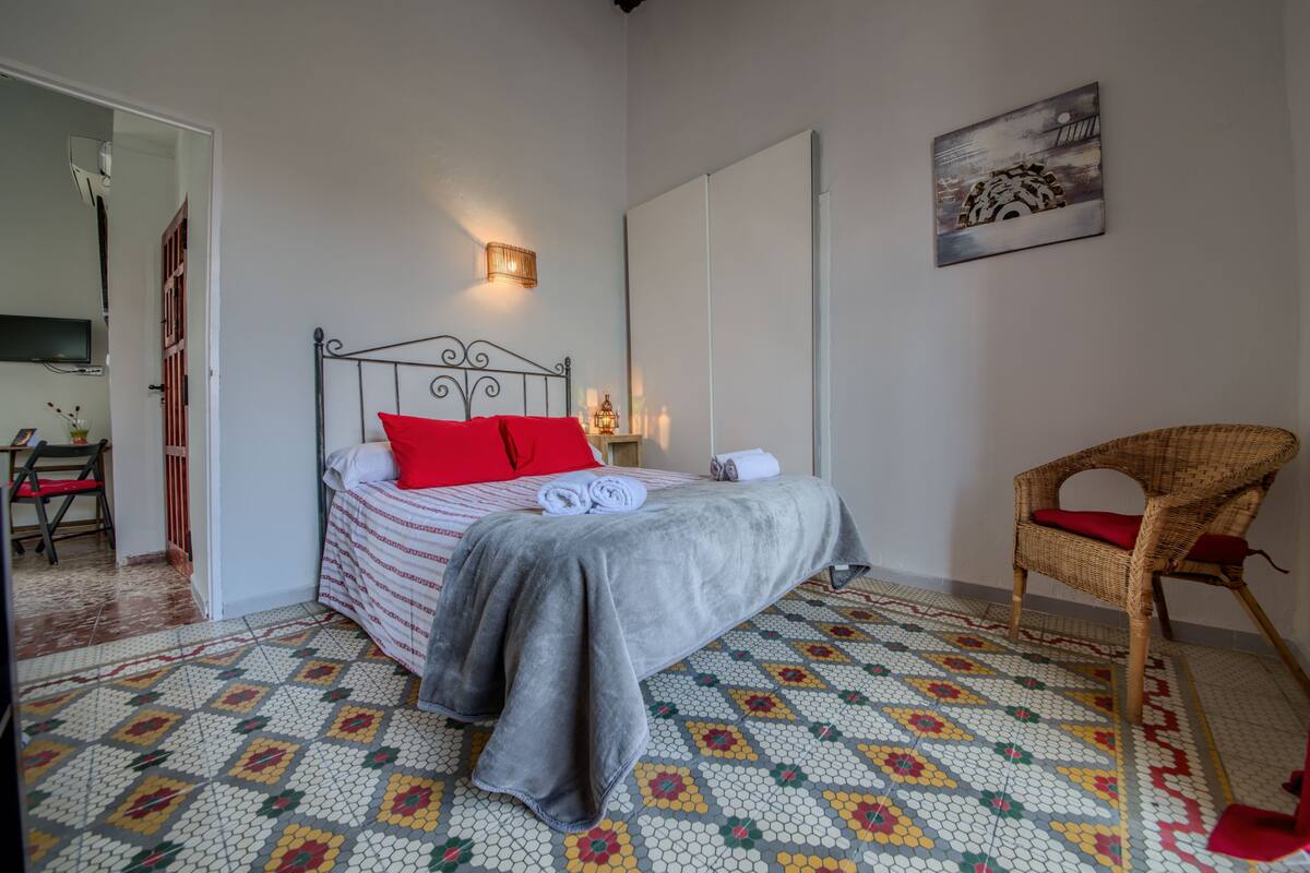 Um quarto com uma cama, uma cadeira, uma luminária na parede e uma porta que dá para outro cômodo.