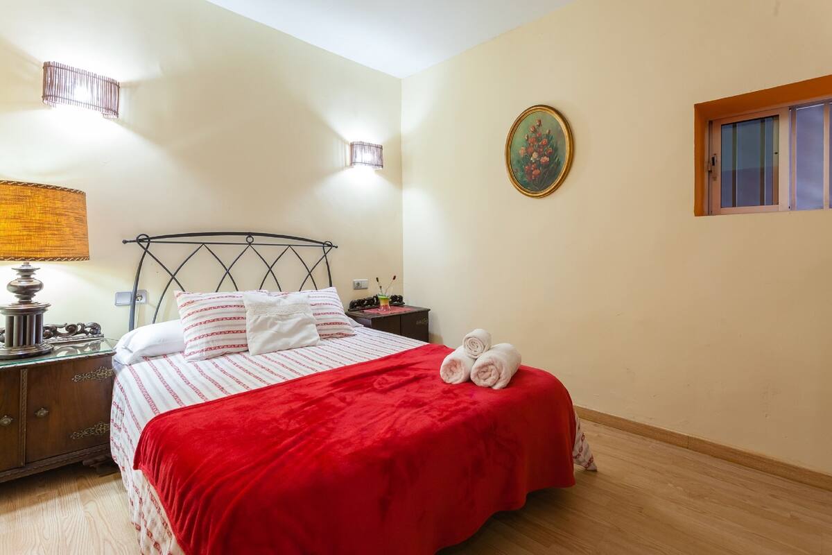 Cama bem arrumada com um cobertor vermelho, mesa de cabeceira de madeira com uma luminária e um quadro na parede.