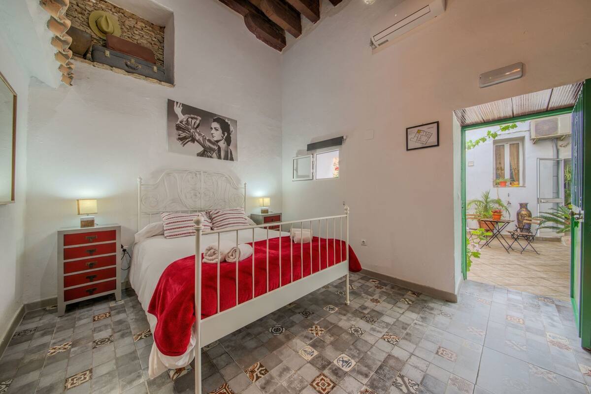 Quarto com cama, mesas de cabeceira, manta vermelha, ladrilhos no chão com padrão e vista para o exterior.