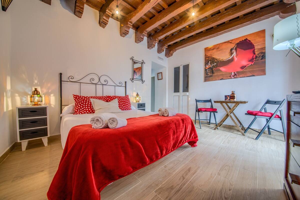 Quarto com colcha vermelha, teto de madeira, quadro na parede e uma pequena mesa com cadeiras.