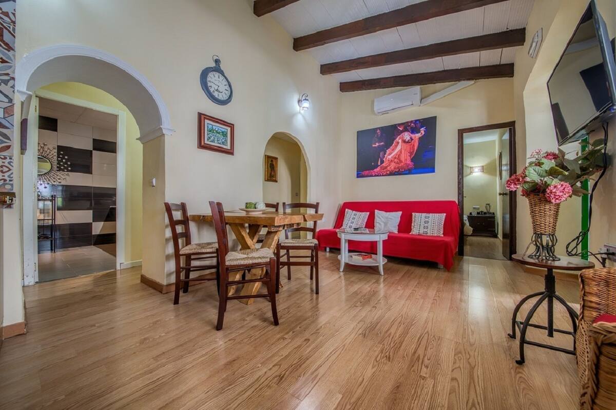 Uma sala de estar ampla com piso de madeira, um sofá vermelho, uma mesa de jantar com cadeiras e um quadro grande na parede.
