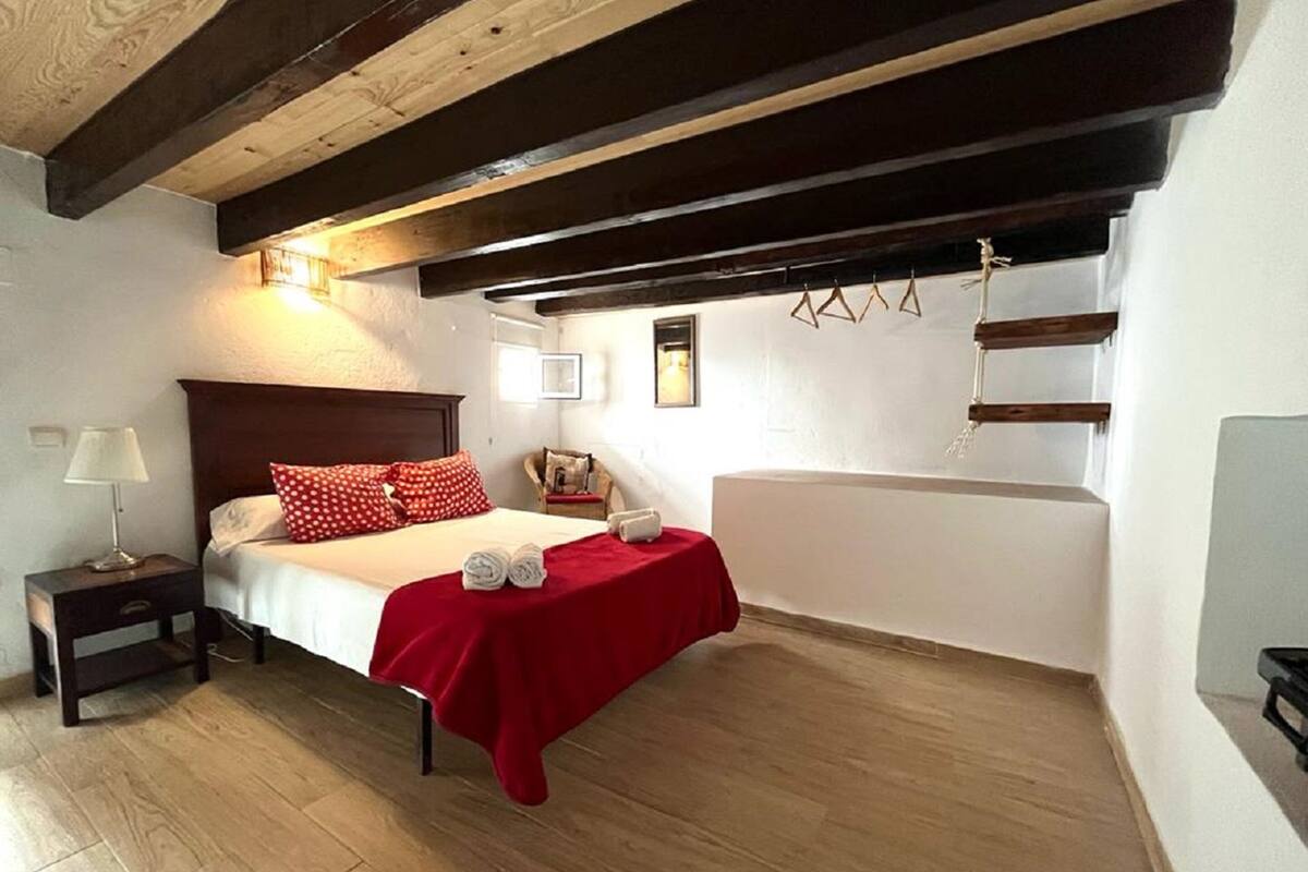 Quarto com teto de madeira, cama com colcha vermelha, mesa de cabeceira com abajur e uma pequena mesa com toalha.