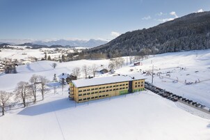 Um edifício grande com telhado plano, rodeado por neve no chão e árvores.