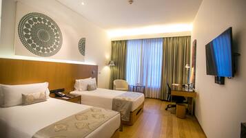 Quarto de hotel com duas camas, uma escrivaninha com cadeira, televisão e uma janela grande com cortinas.