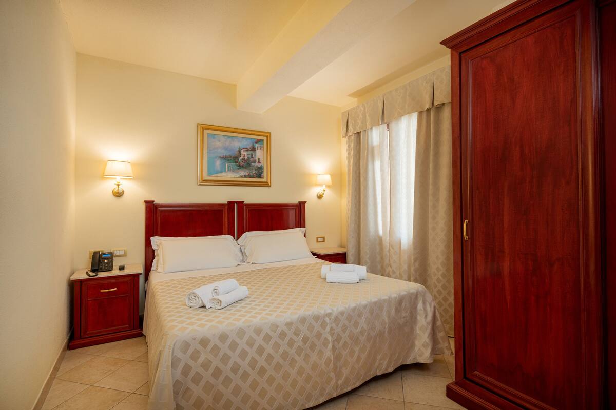 Quarto de hotel com uma cama grande, duas mesas de cabeceira, um guarda-roupa de madeira, um quadro na parede e duas luminárias de parede.