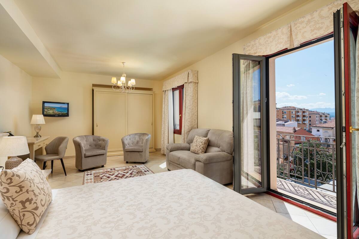 Quarto de hotel com cama, duas poltronas, escrivaninha com abajur, candelabro e varanda com vista para edifícios.