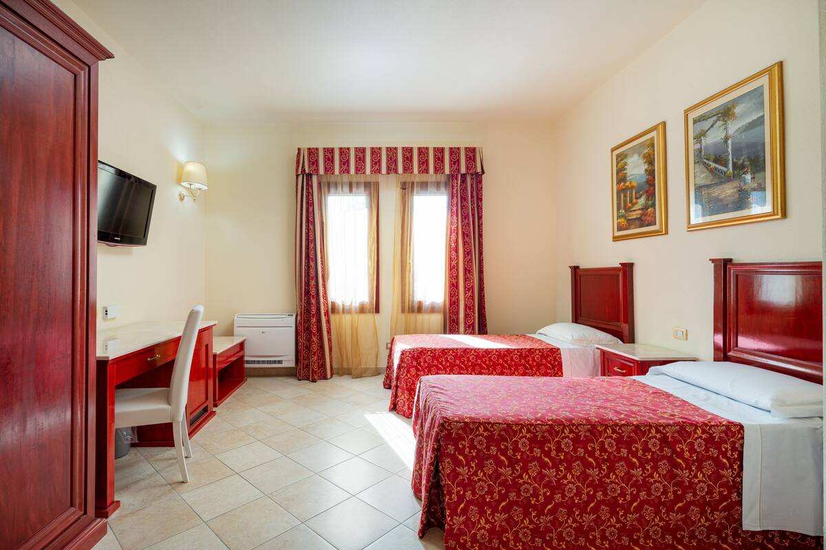 Quarto de hotel com duas camas, uma mesa, uma cadeira, uma televisão e duas pinturas na parede.