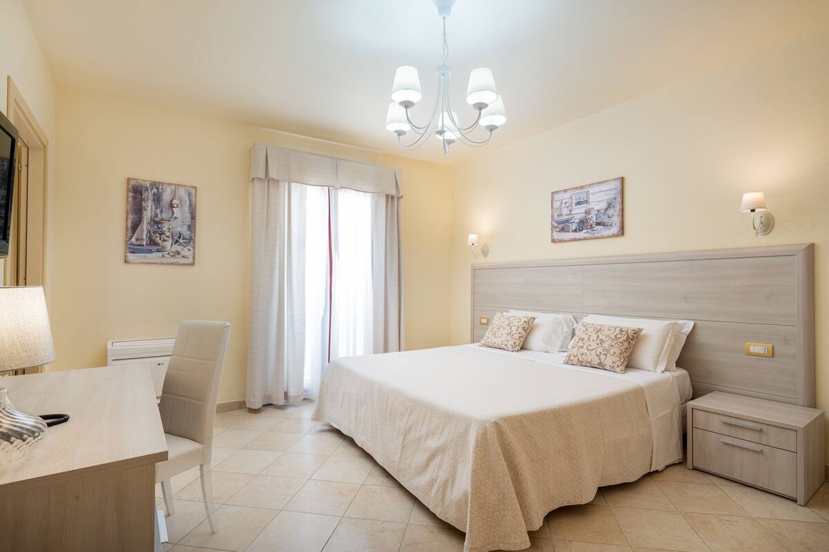 Quarto de hotel com uma cama grande, mesas de cabeceira, escrivaninha com cadeira, lustre e obras de arte nas paredes.