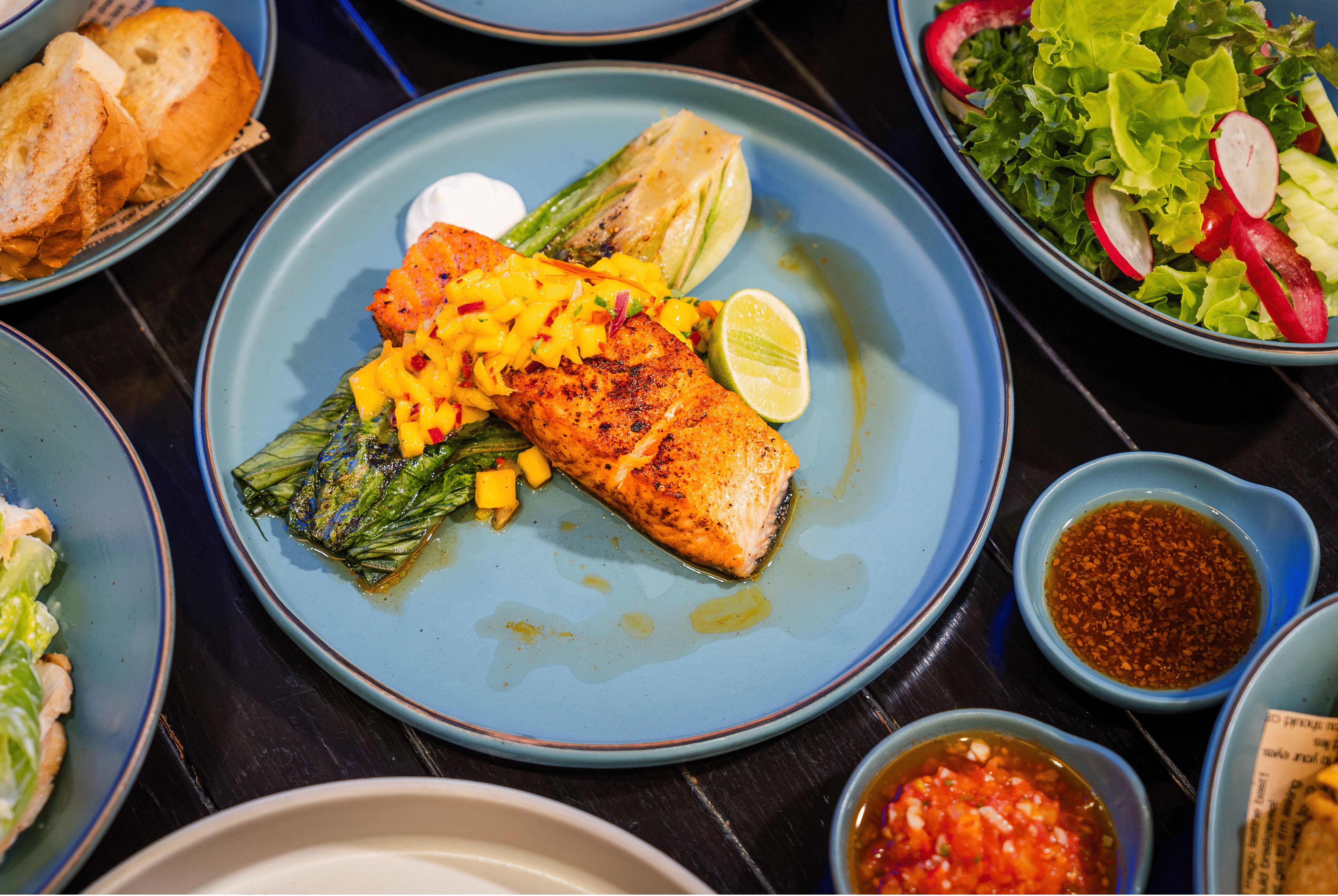 Diversos pratos sobre uma mesa, incluindo salmão grelhado com salsa de manga, aspargos e uma salada.