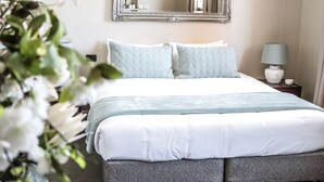 Cama bem arrumada com colcha branca e azul clara, cabeceira estofada cinza e mesa de cabeceira com abajur.