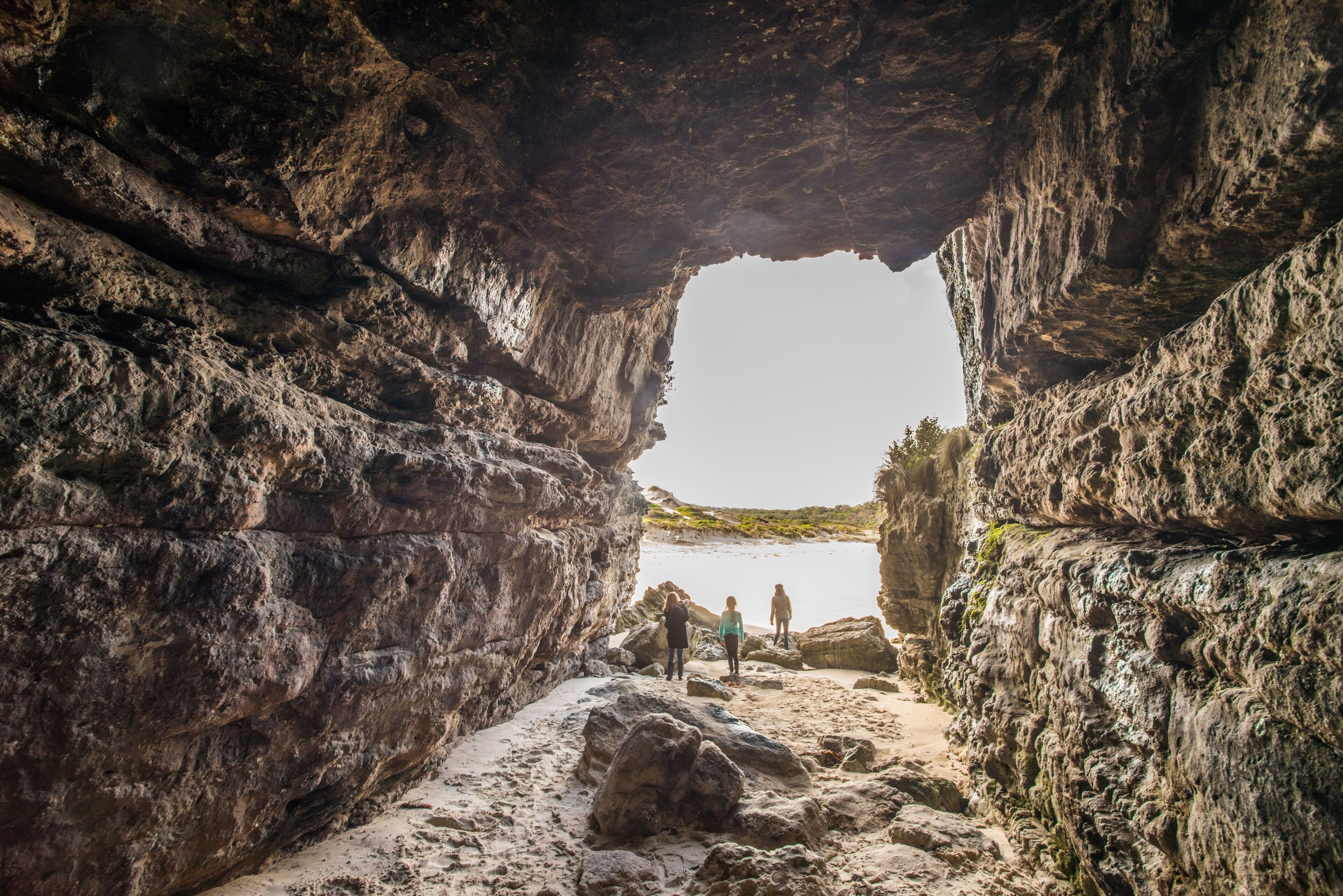 Entrada rochosa de uma caverna com vista para a praia e pessoas na abertura.