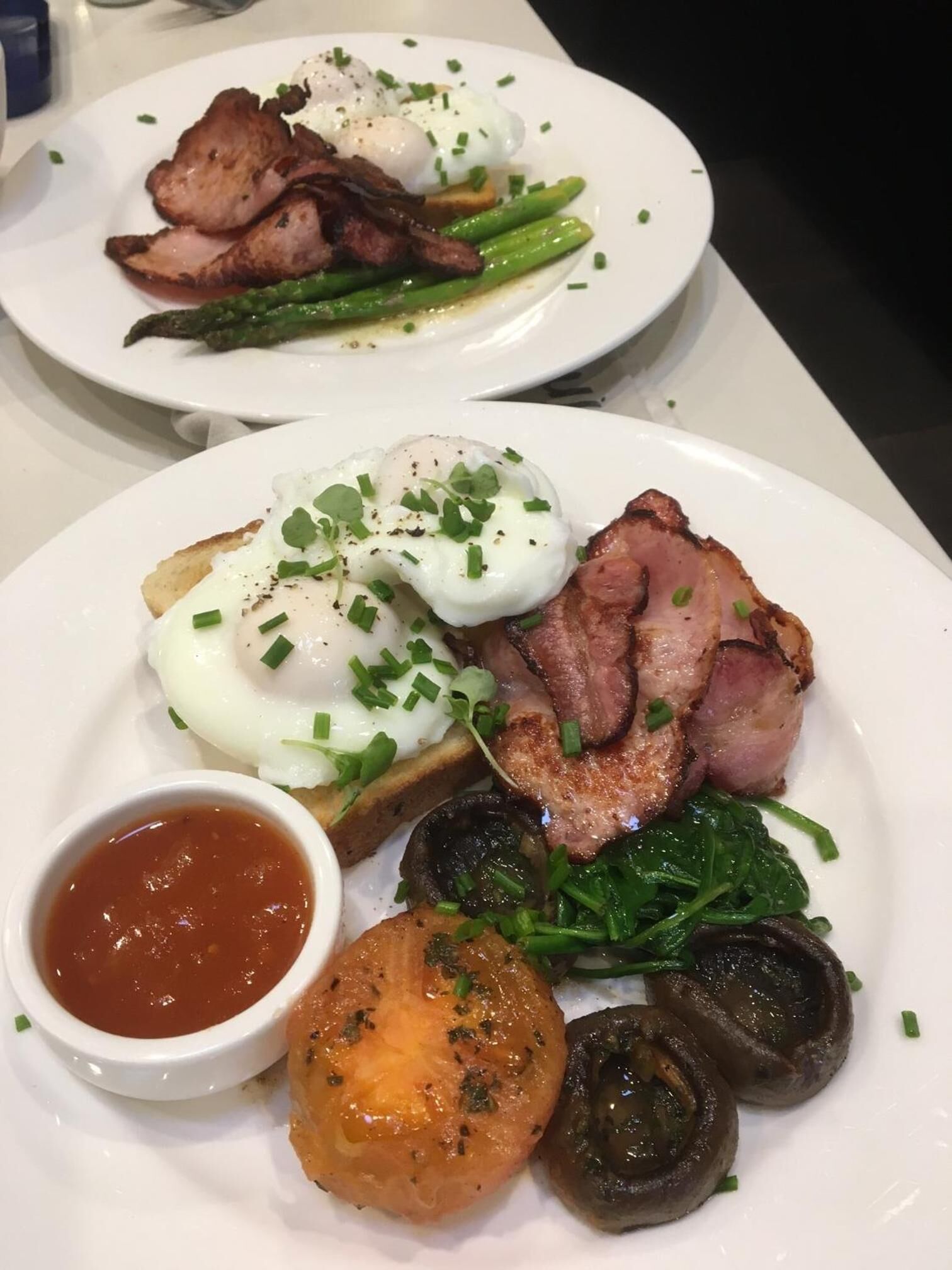 Prato de café da manhã com bacon, ovos, aspargos, cogumelos e uma pequena tigela de molho.