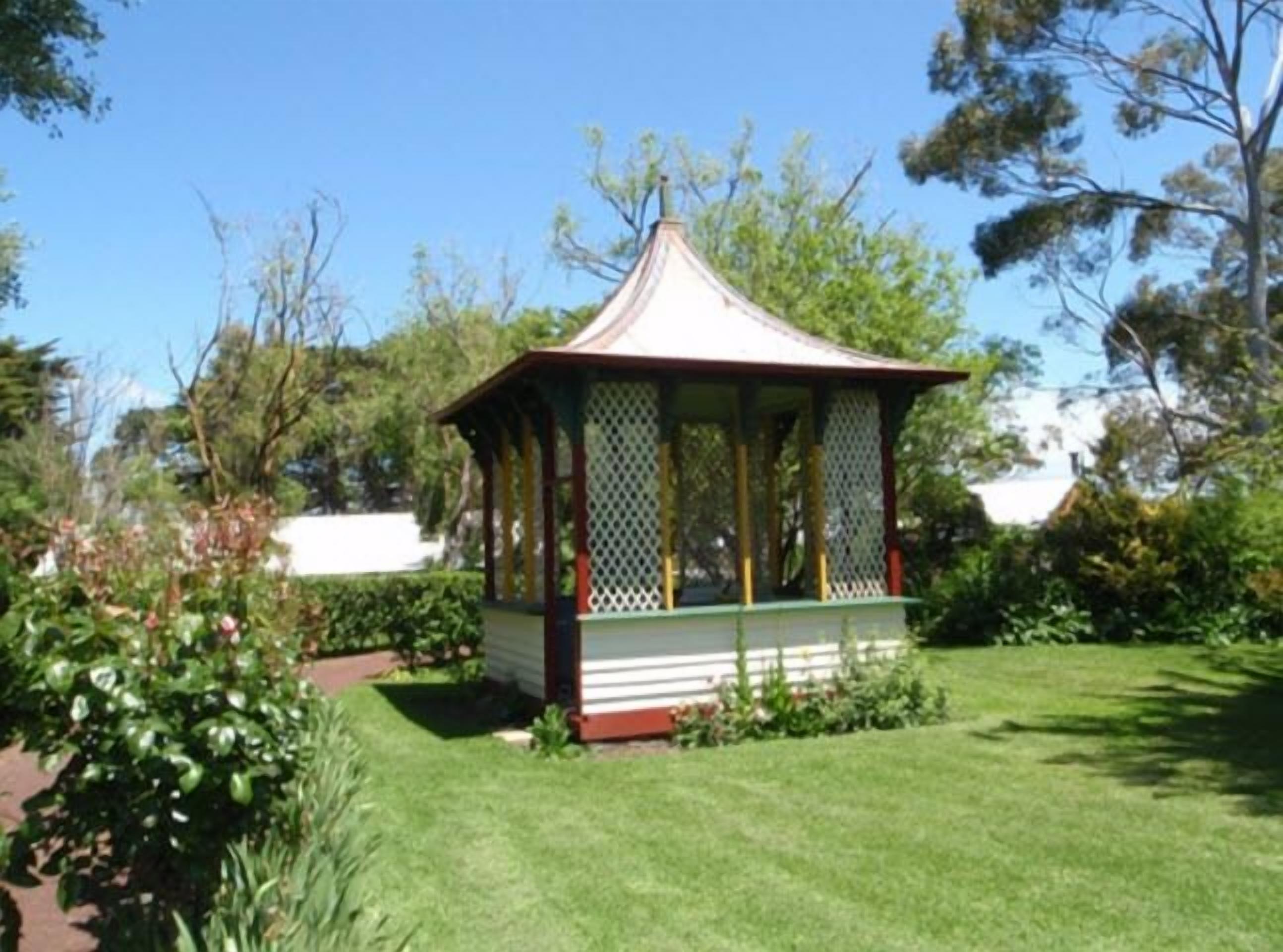 Um gazebo com teto treliçado e base verde, cercado por gramado e arbustos bem cuidados.