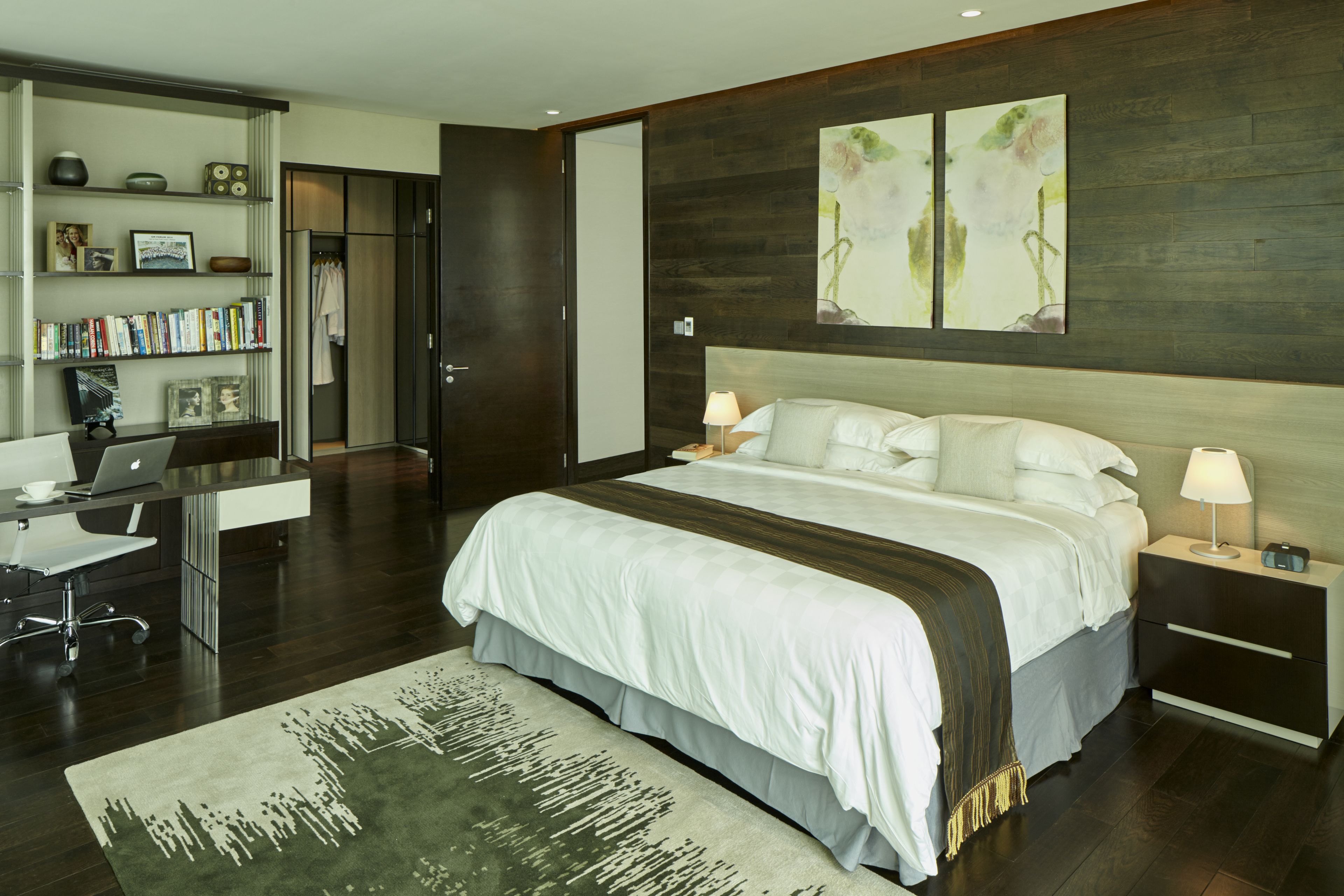 Quarto de hotel moderno com uma cama grande, uma escrivaninha com um laptop, uma estante de livros e um quadro na parede.