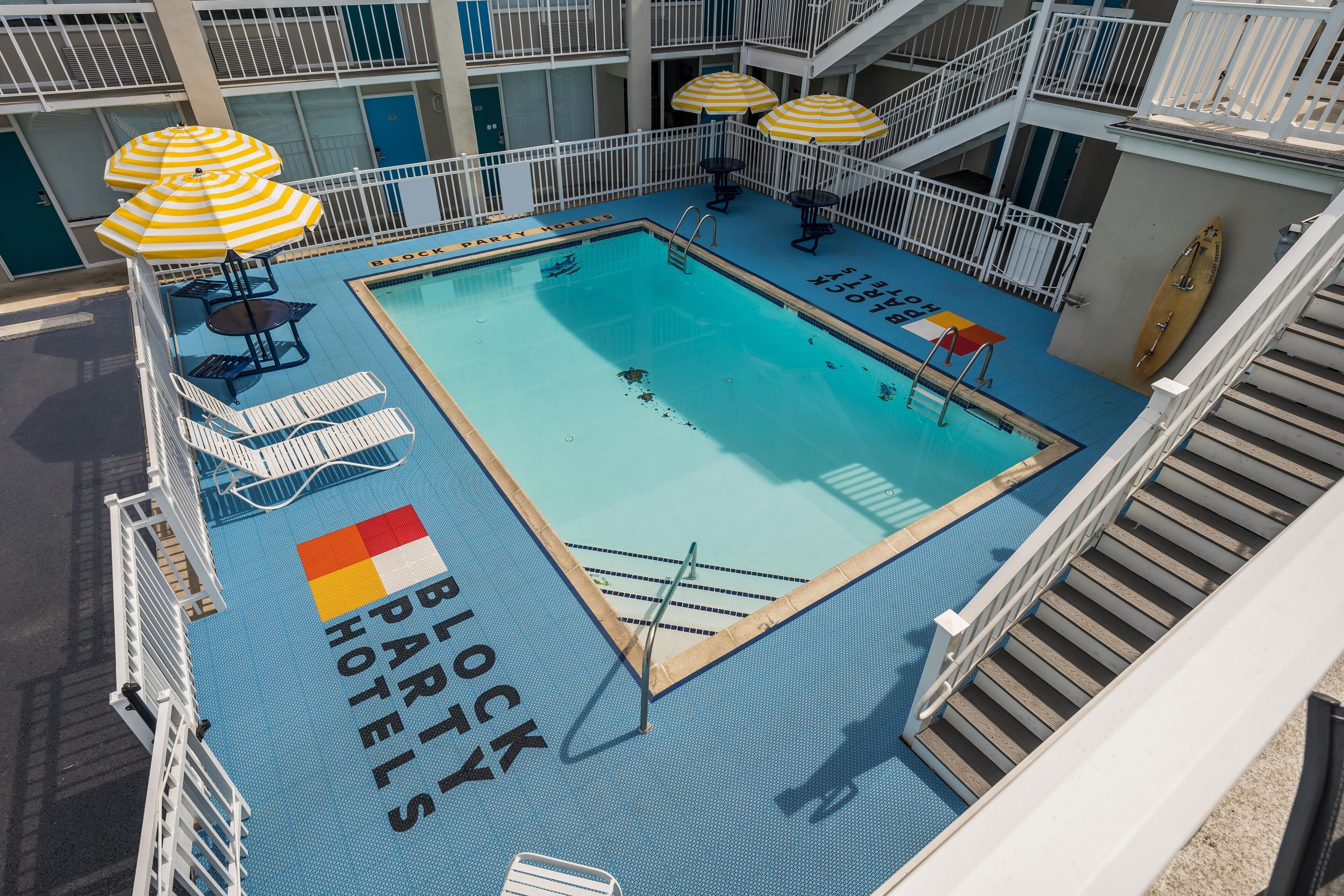 Área da piscina com espreguiçadeiras e guarda-sóis, posto de salva-vidas e uma prancha de surf.