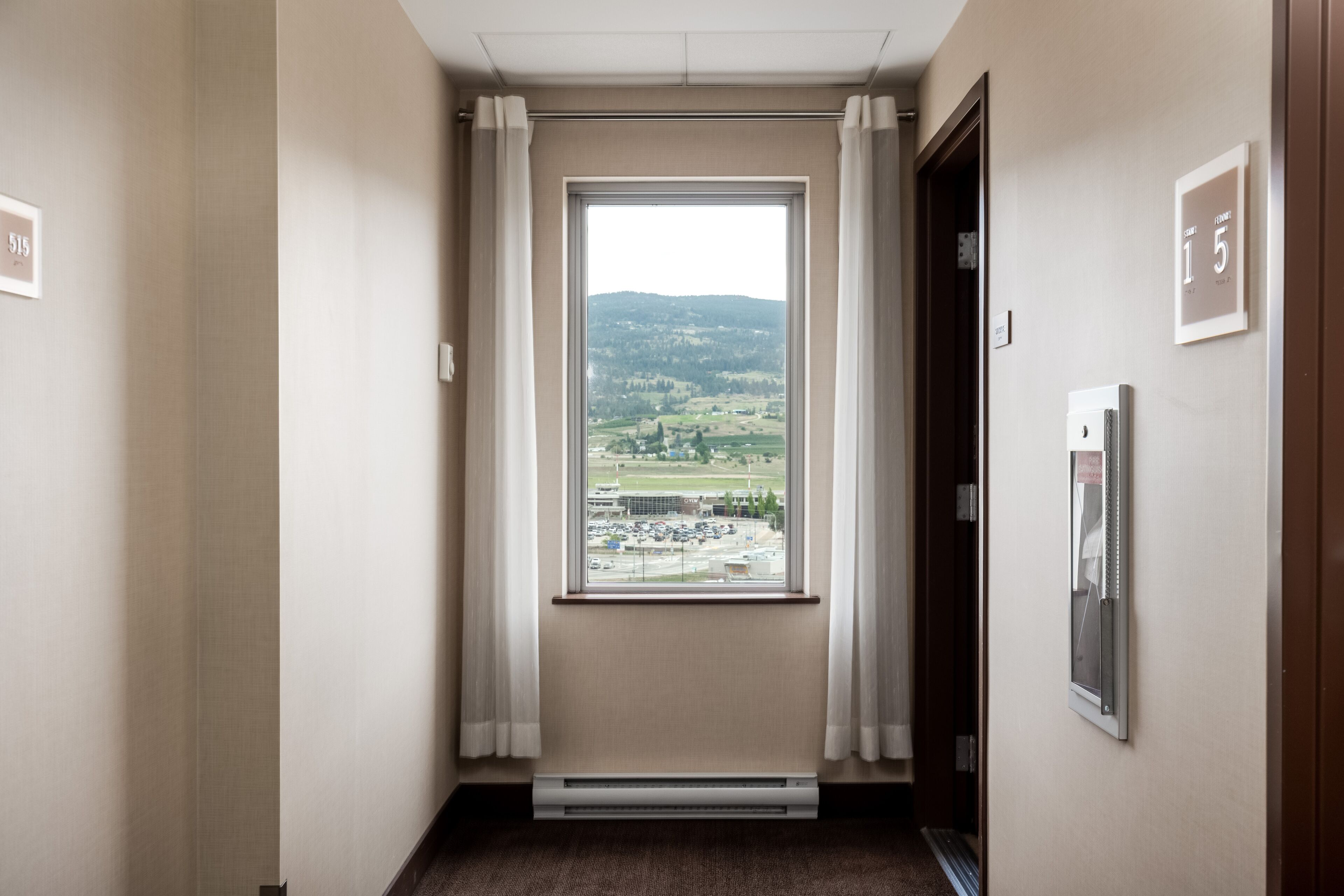 Corredor de hotel com uma janela grande que oferece vista para um estacionamento e colinas ao longe.
