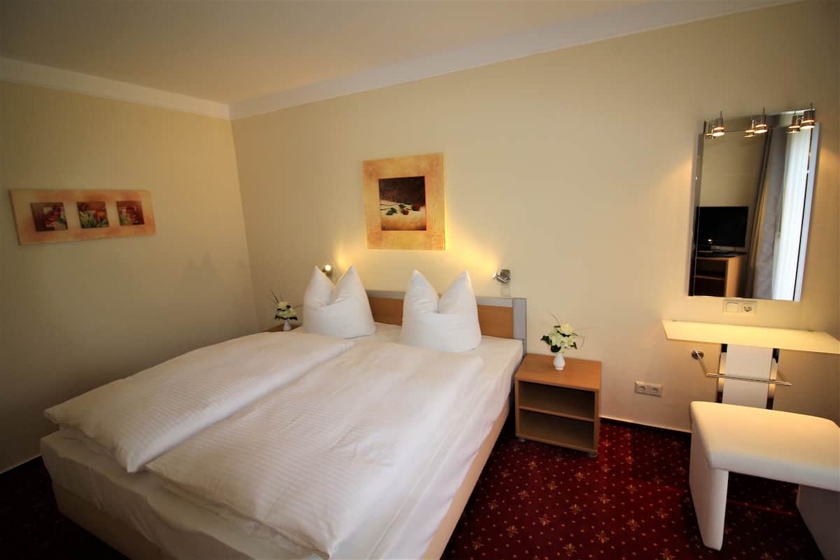 Quarto de hotel com uma cama grande, mesa de cabeceira com abajur, pequena escrivaninha, cadeira, espelho e obras de arte nas paredes.