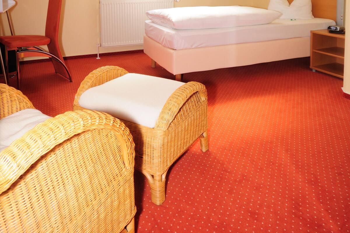 Quarto com cama, duas cadeiras de vime, uma mesinha, radiador e janela com cortina.