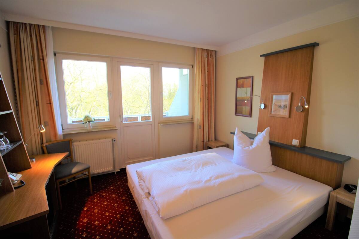 Quarto de hotel com uma cama grande, uma escrivaninha e uma cadeira. Há uma janela com cortinas, um radiador e uma cabeceira de madeira.