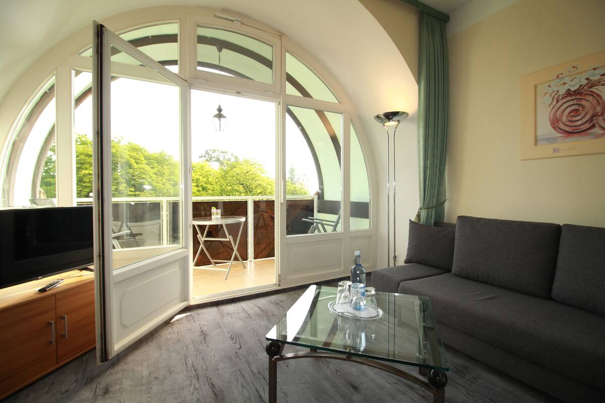 Uma sala de estar moderna com uma mesa de centro de vidro, um sofá cinzento e uma TV sobre um armário de madeira. Uma grande janela em arco oferece vista para o exterior.