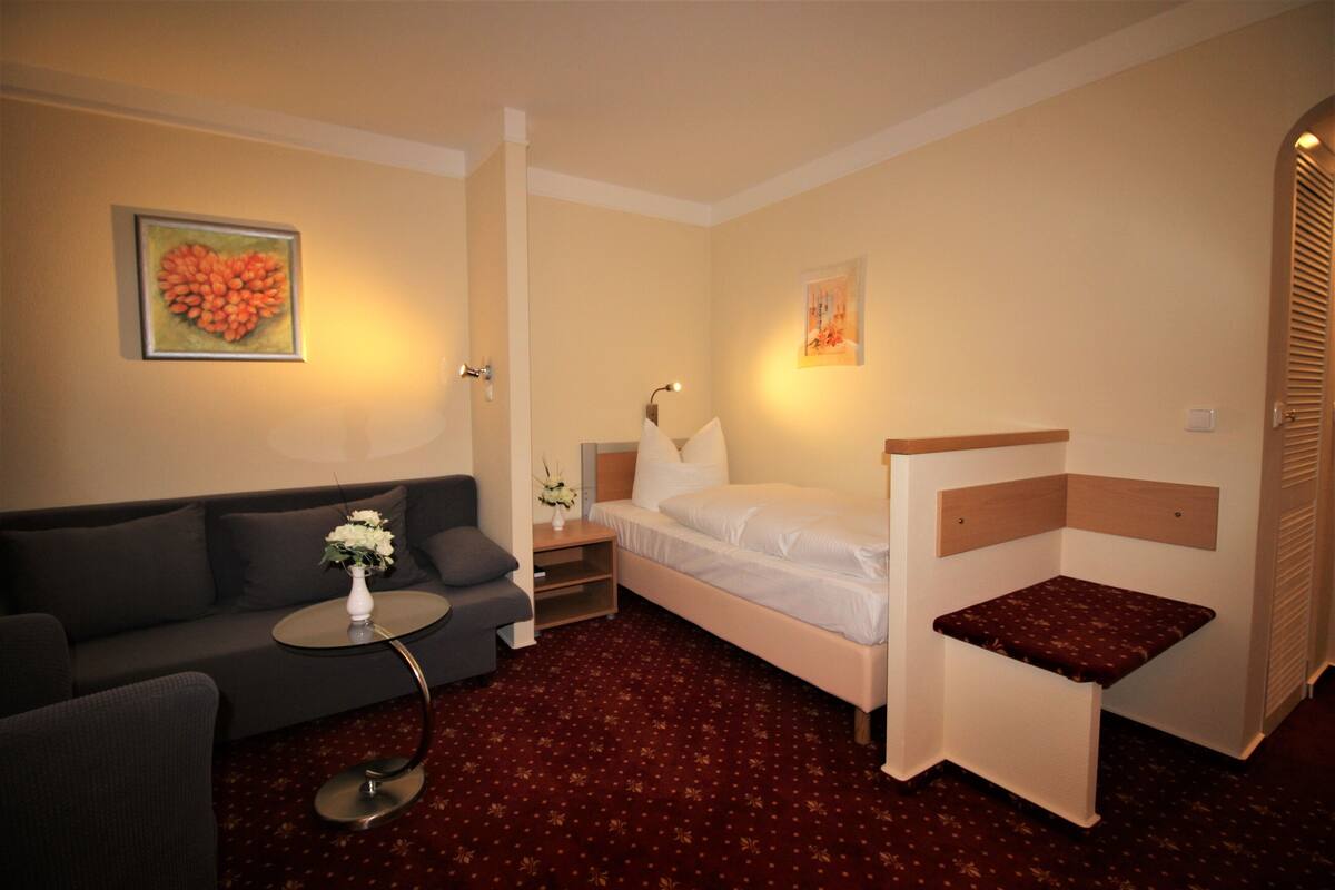 Quarto de hotel compacto com uma cama, um sofá, uma pequena mesa com um vaso e uma obra de arte pendurada na parede.