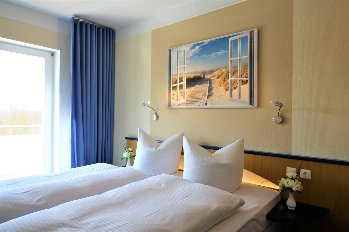 Quarto de hotel com uma cama grande, uma janela com cortinas azuis, um quadro pendurado na parede de uma praia e uma mesa de cabeceira com um vaso de flores.