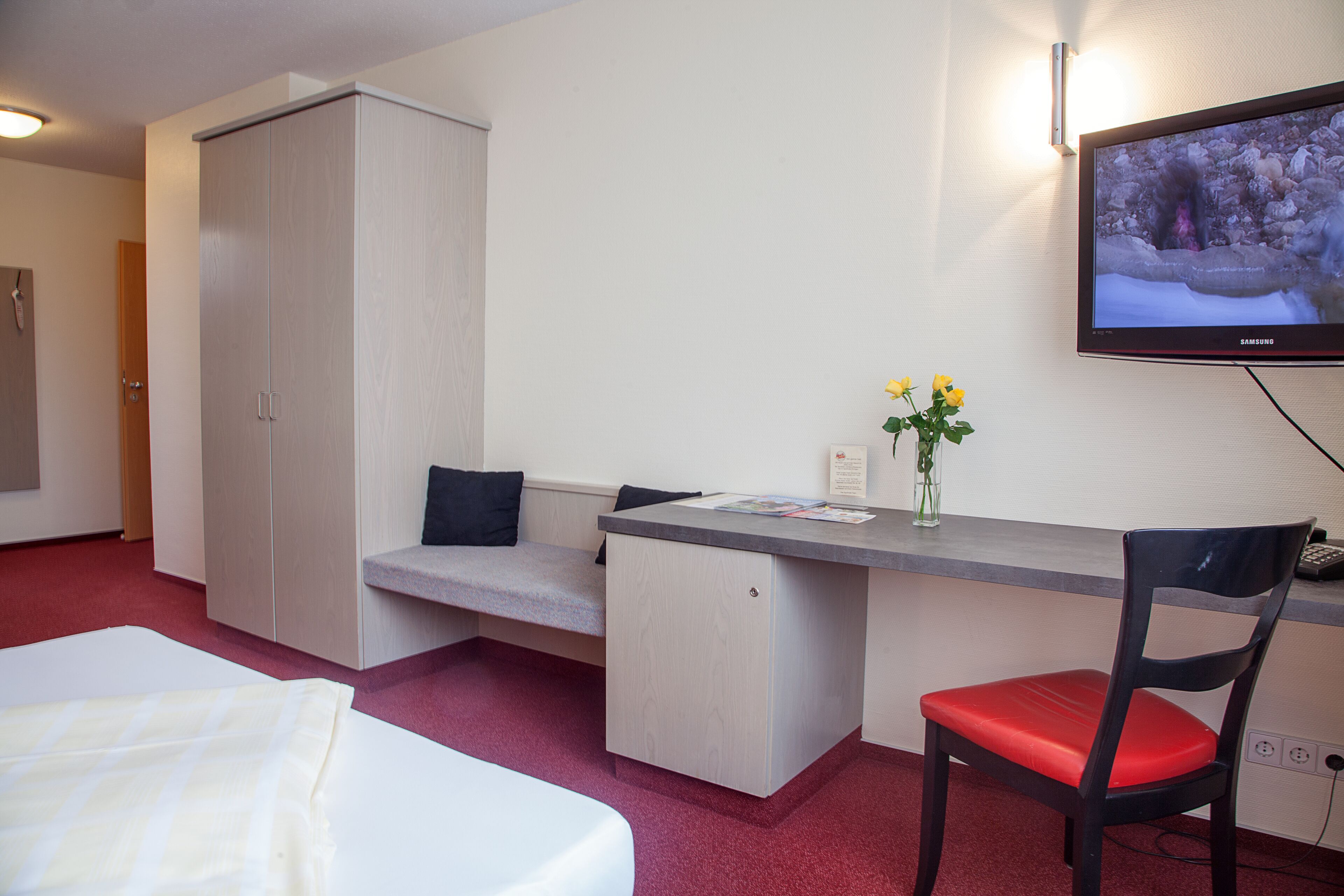 Quarto de hotel com cama, escrivaninha com cadeira, televisão e guarda-roupa.