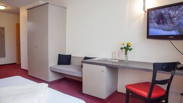 Quarto de hotel com cama, escrivaninha com cadeira, televisão e guarda-roupa.