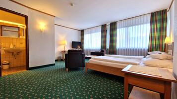 Quarto de hotel com cama, secretária, cadeira, televisão, banheiro e janela com cortinas.