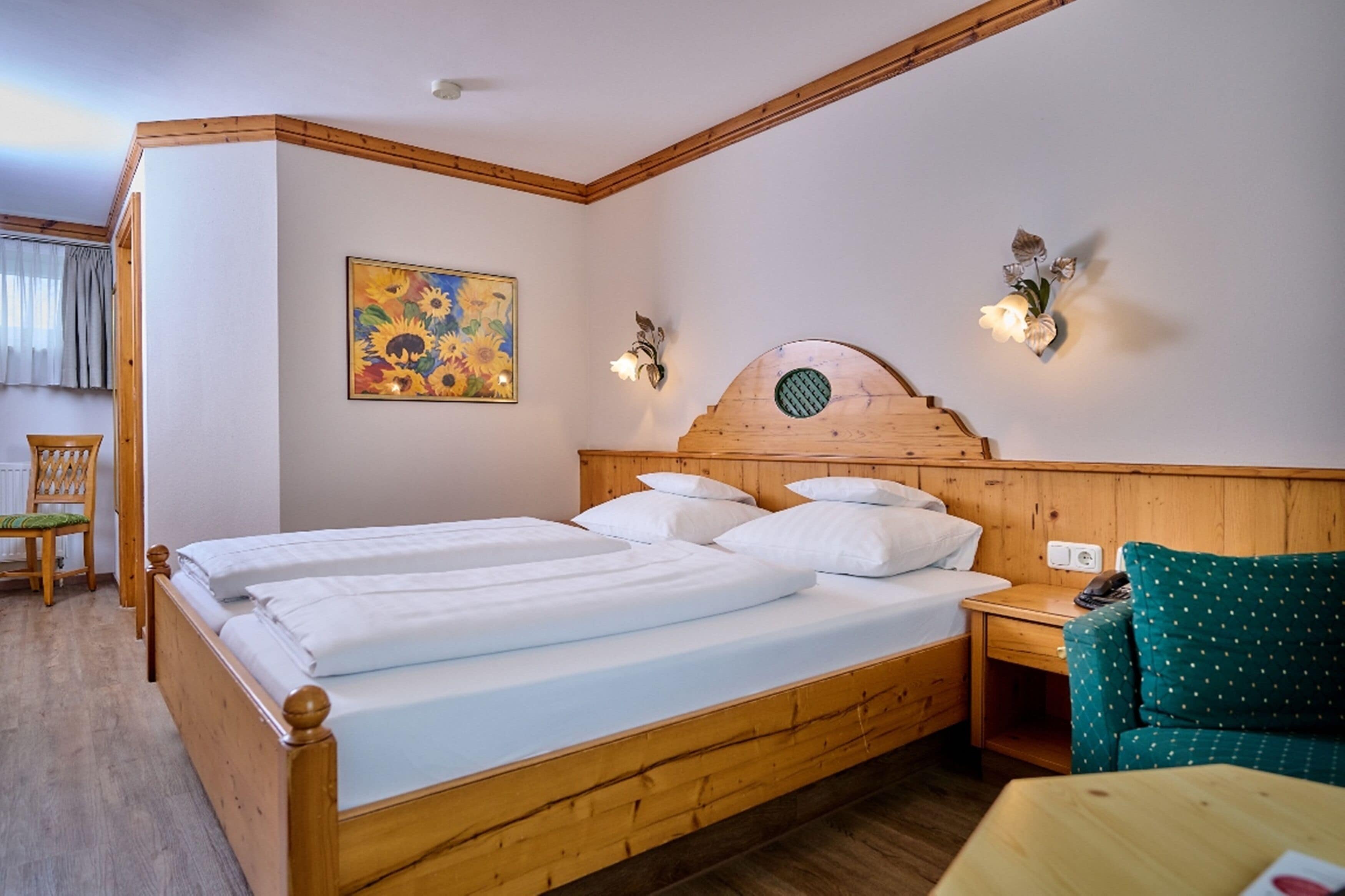 Cama de casal com cabeceira de madeira, duas mesas de cabeceira, uma cadeira verde com padrão e um aplique de parede.