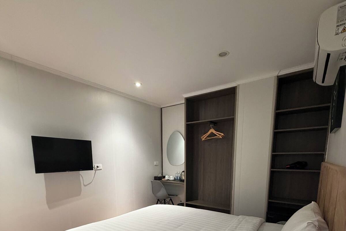 Quarto de hotel com cama, televisão presa na parede, espelho, guarda-roupa e prateleira com artigos de higiene.