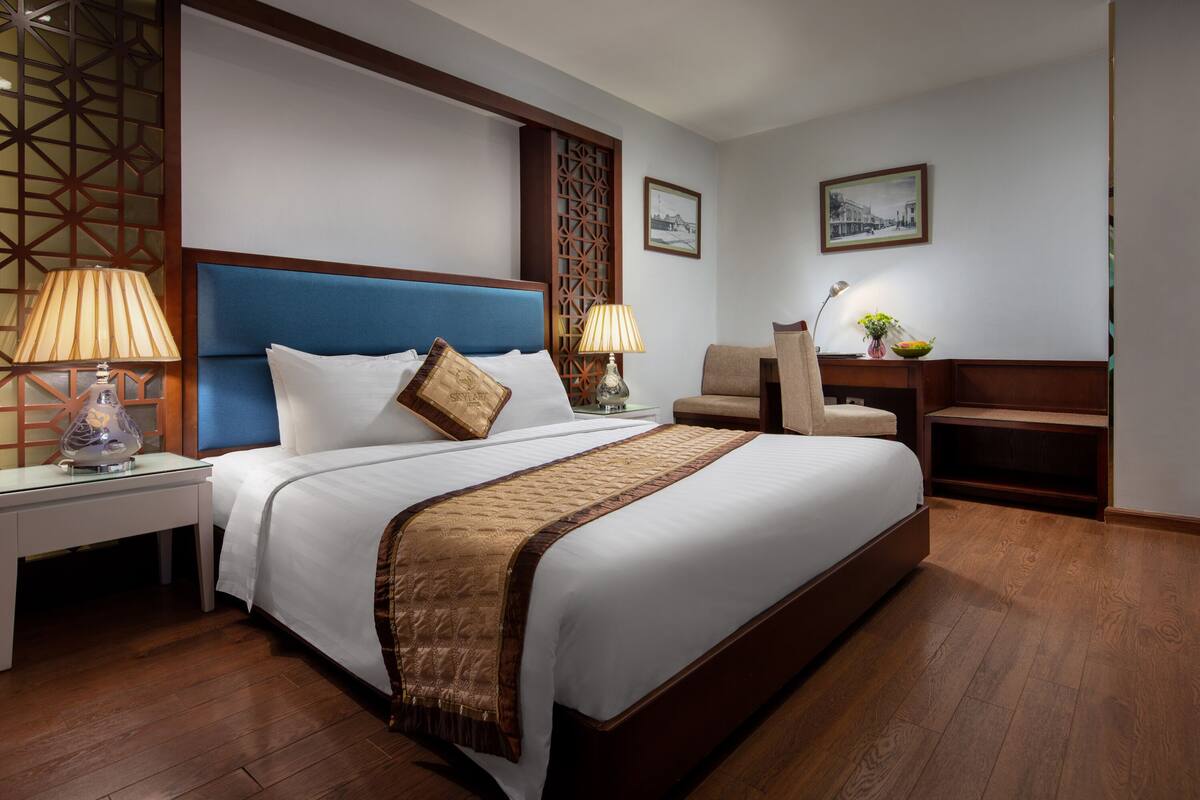Quarto de hotel com uma cama grande, duas mesas de cabeceira, um criado-mudo, uma cabeceira de madeira, um painel decorativo na parede, uma cadeira, uma pequena mesa com um vaso e duas molduras com quadros.